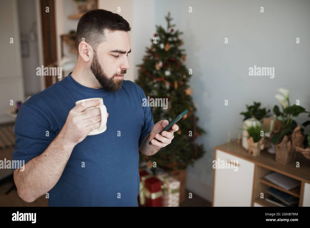 Serious young Caucasian man with beard standing with mug in living room ...
