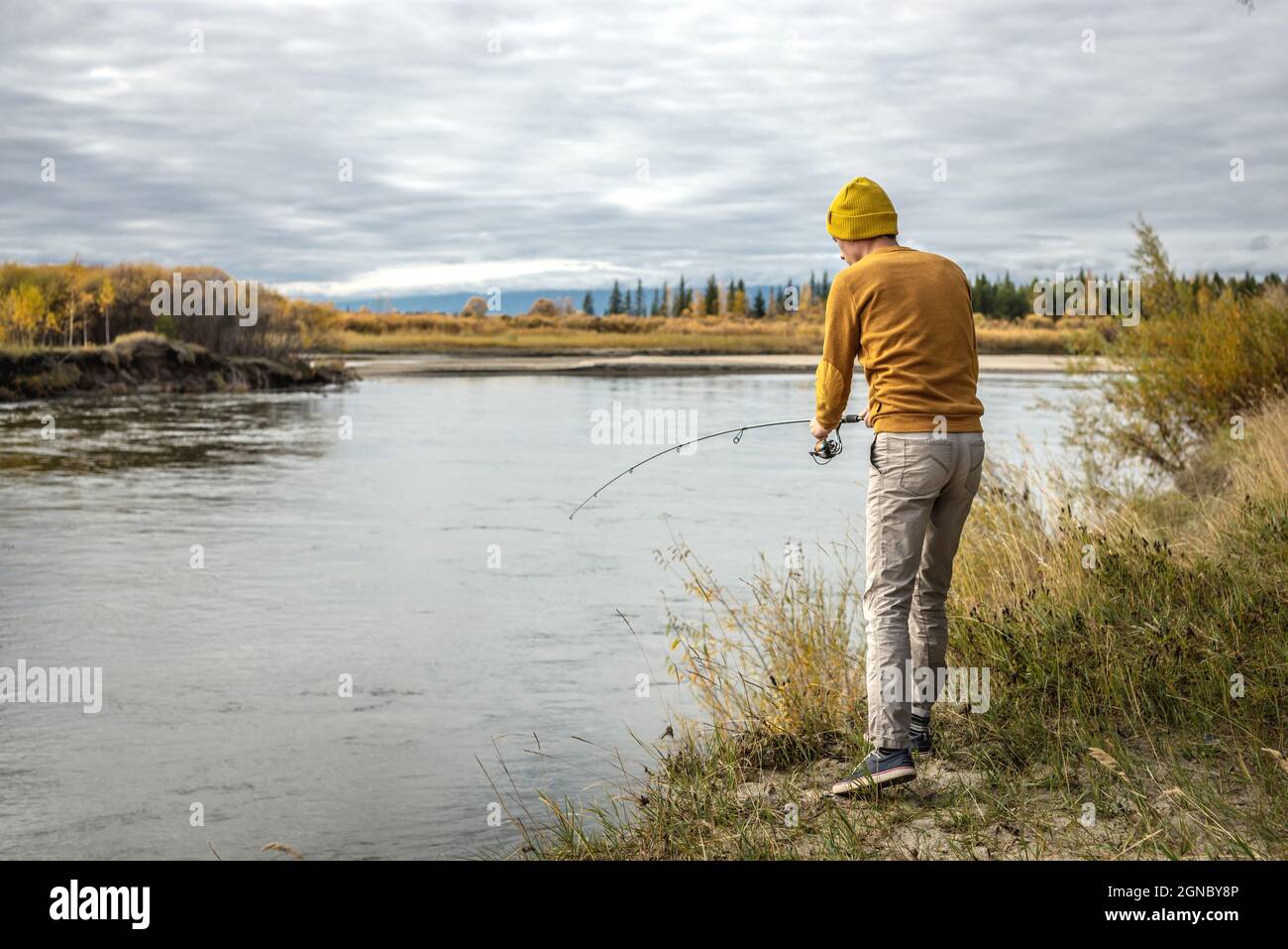 Young man fisherman in a yellow sweater and hat is fishing on the river ...
