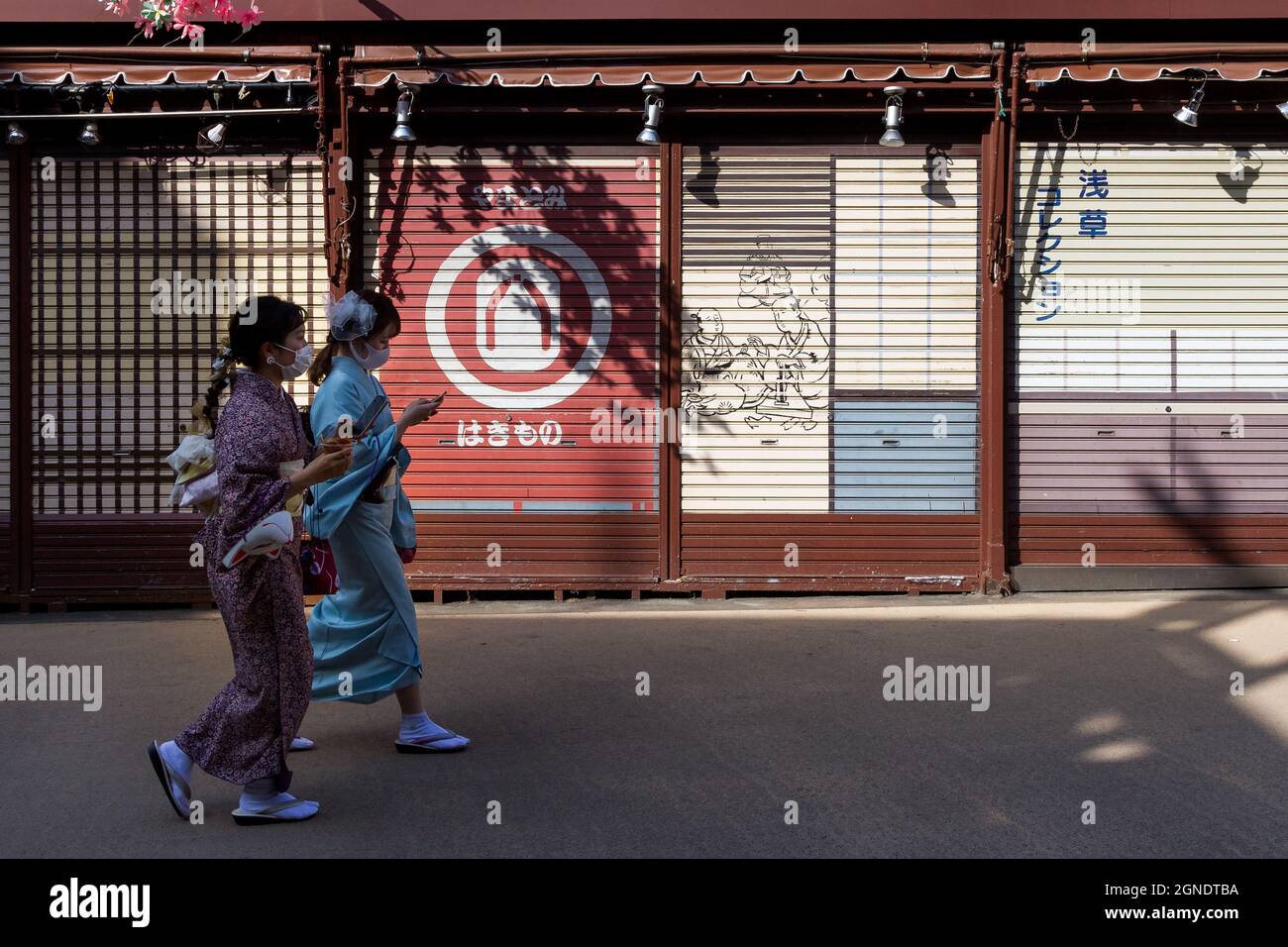 Two young women, wearing kimono and face masks , walk past shuttered ...