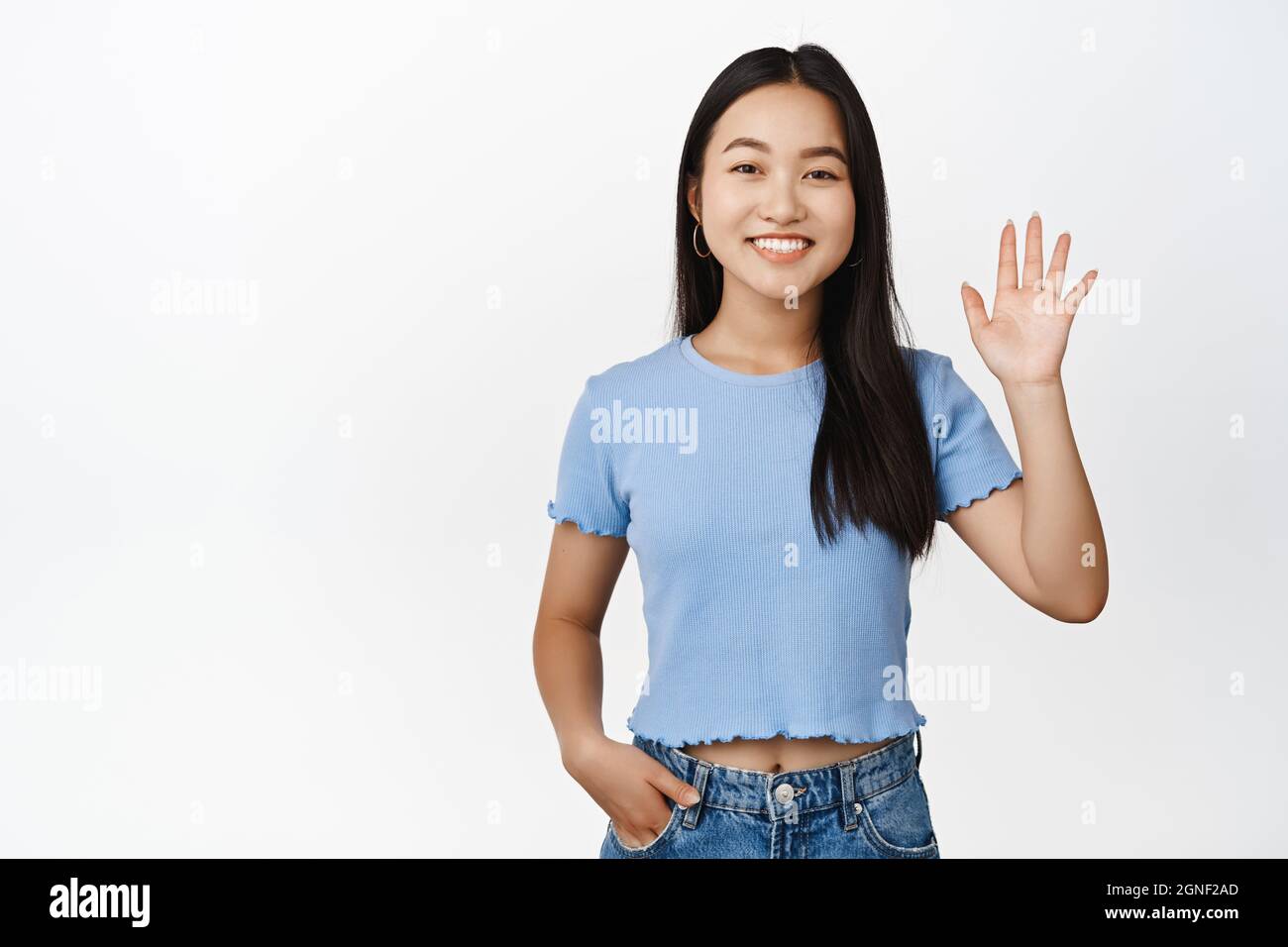 Happy asian girl saying hello, smiling and waving hand to greet you ...