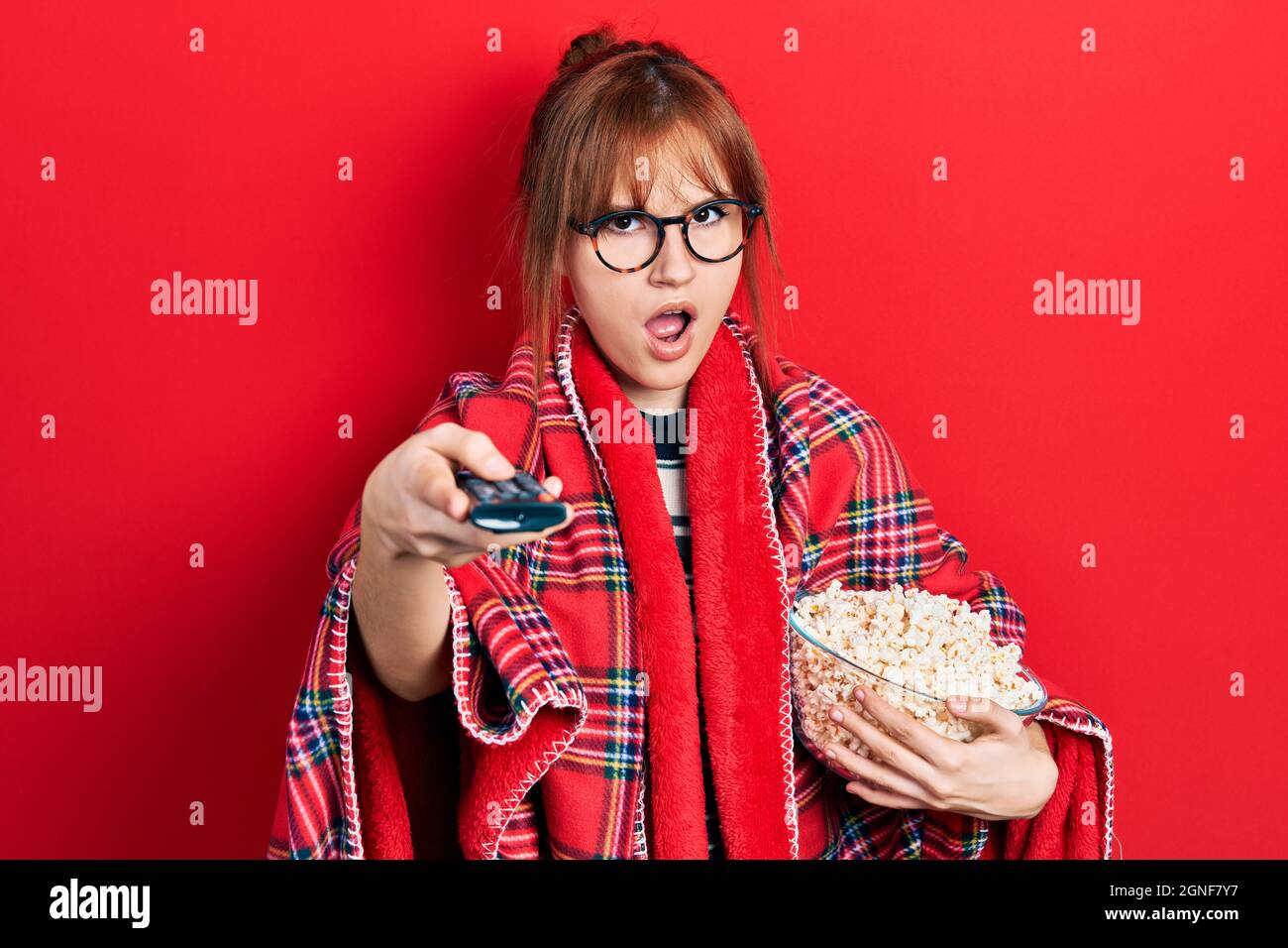 Redhead young woman eating popcorn using tv control in shock face ...