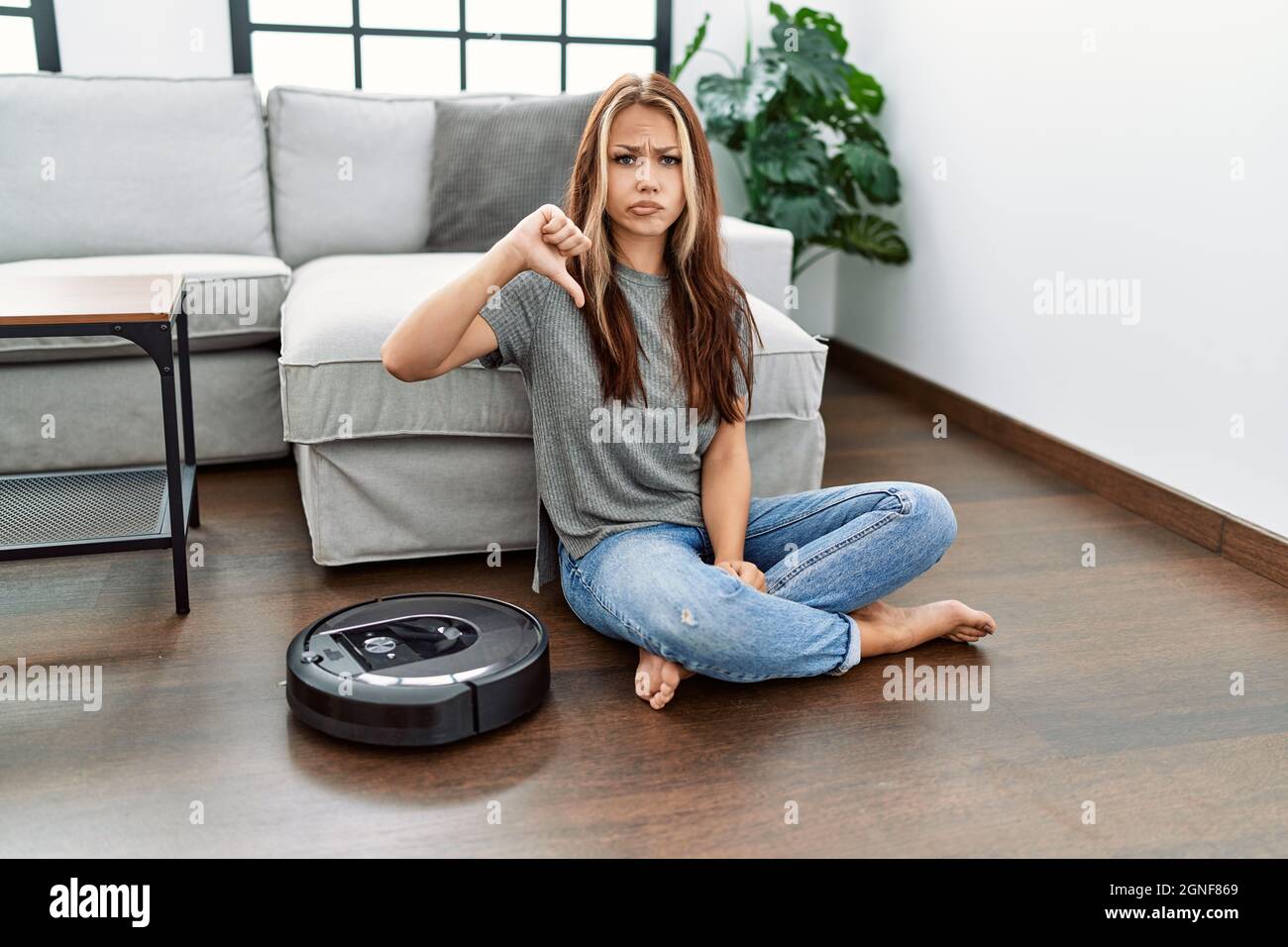 Young caucasian woman sitting at home by vacuum robot with angry face ...