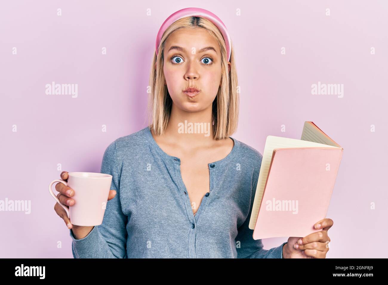Beautiful blonde woman reading a book and drinking a cup of coffee puffing cheeks with funny ...