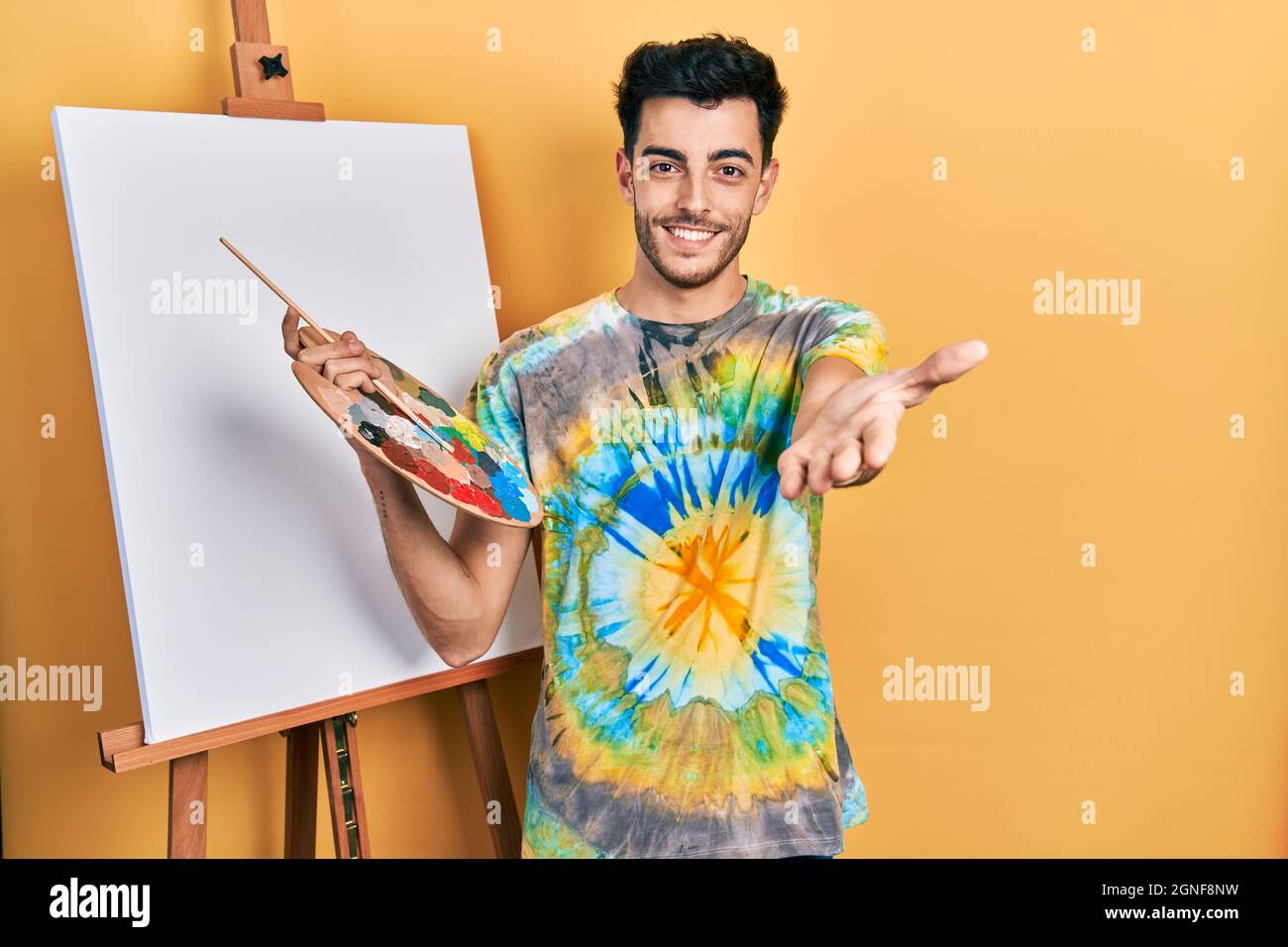 Young hispanic man standing drawing with palette by painter easel stand ...
