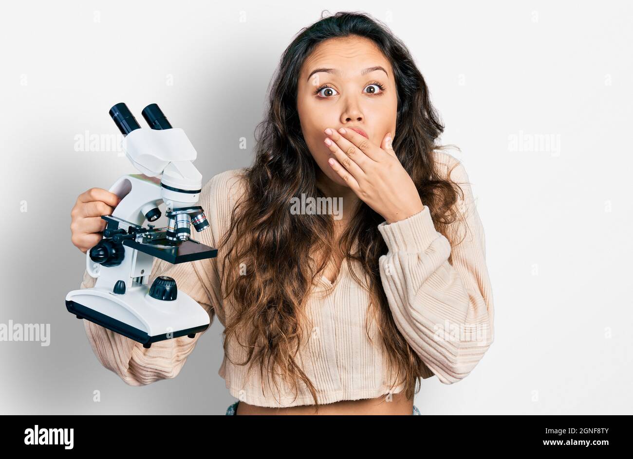 Young hispanic girl holding microscope covering mouth with hand ...