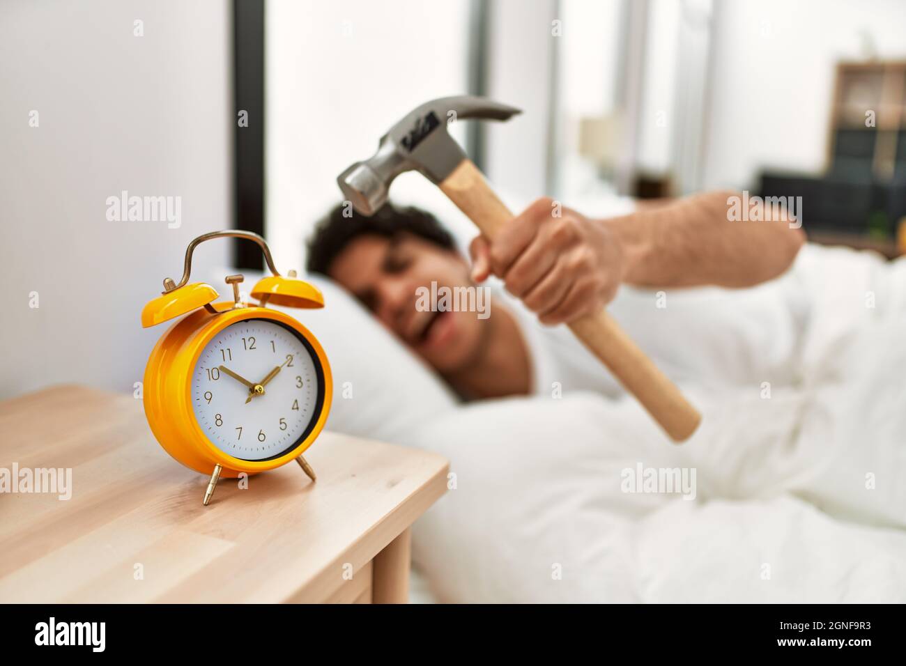 Young Hispanic Man Turning Off Alarm Clock Using Hammer Lying On The