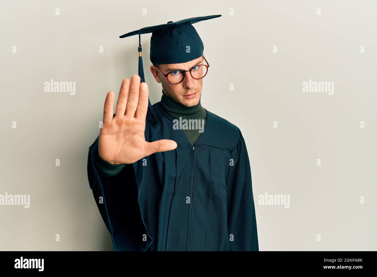 Young caucasian man wearing graduation cap and ceremony robe doing stop ...