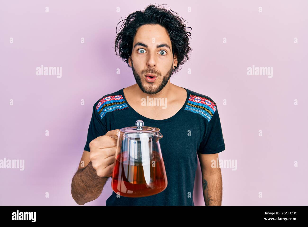Handsome hispanic man holding traditional tea pot scared and amazed ...
