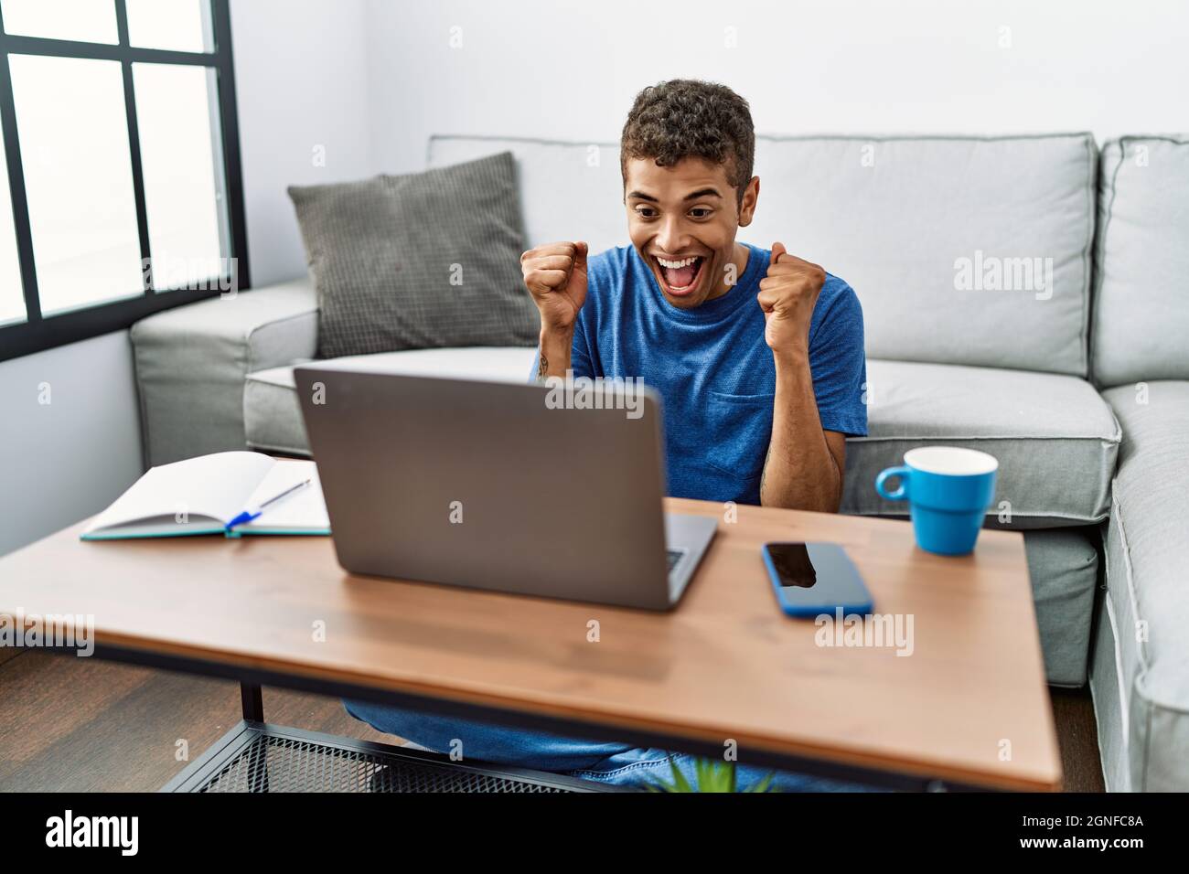 Young handsome hispanic man using laptop sitting on the floor excited ...