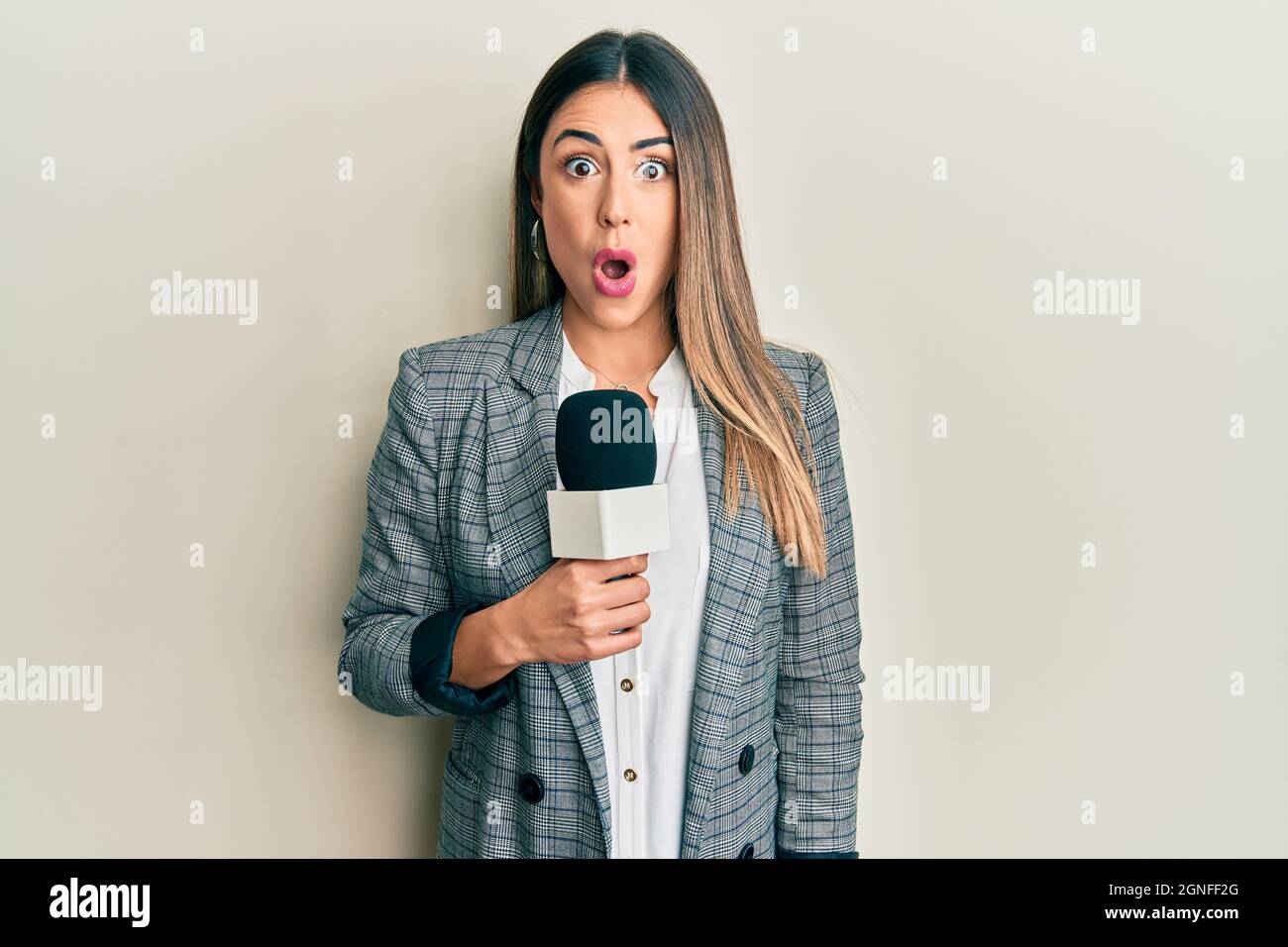 Young hispanic woman holding reporter microphone scared and amazed with ...
