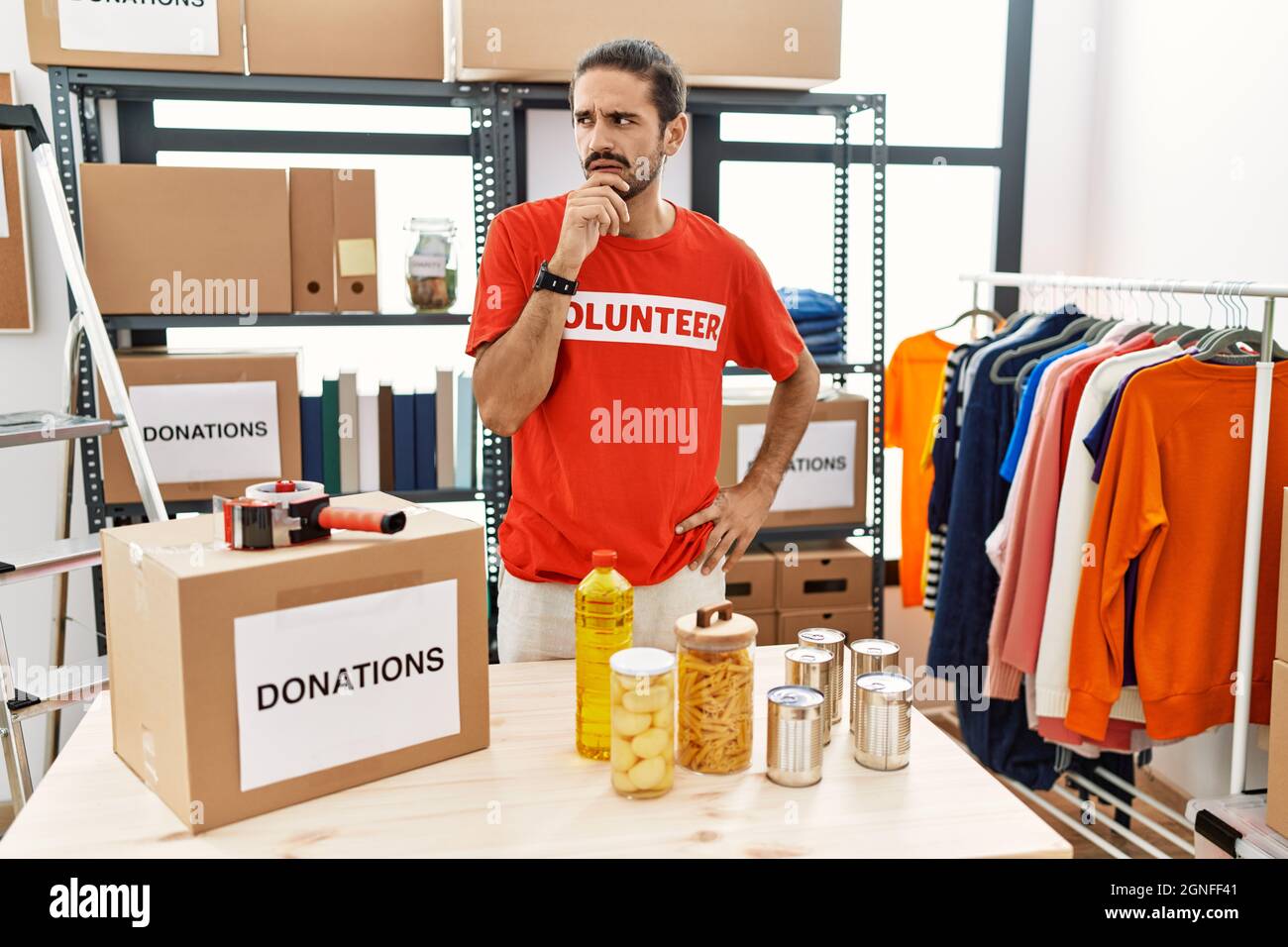 Young hispanic man wearing volunteer t shirt at donations stand looking ...