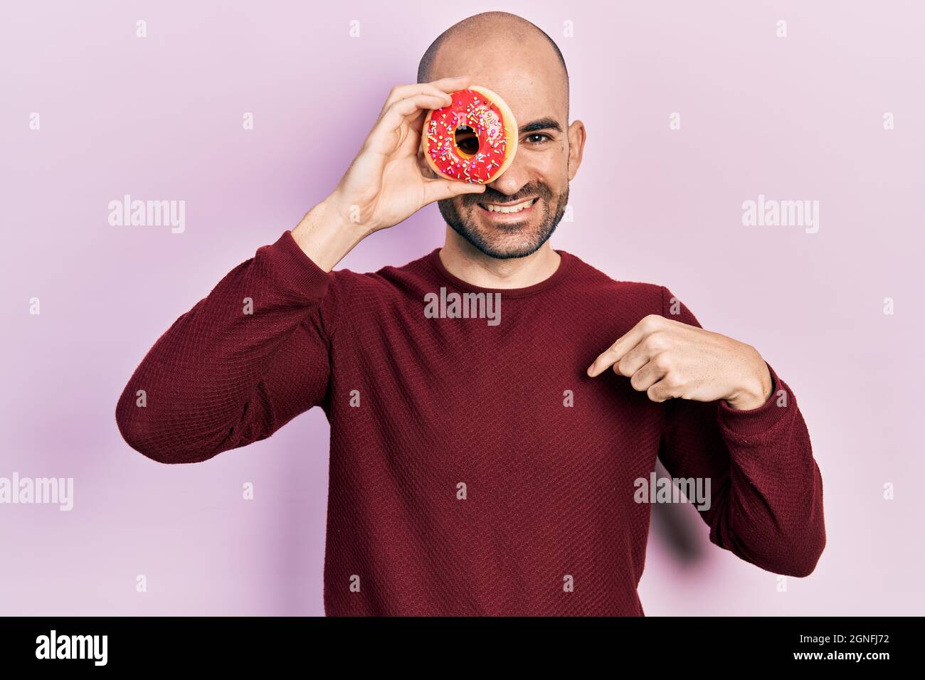 Young bald man holding tasty colorful doughnut on eye pointing finger ...