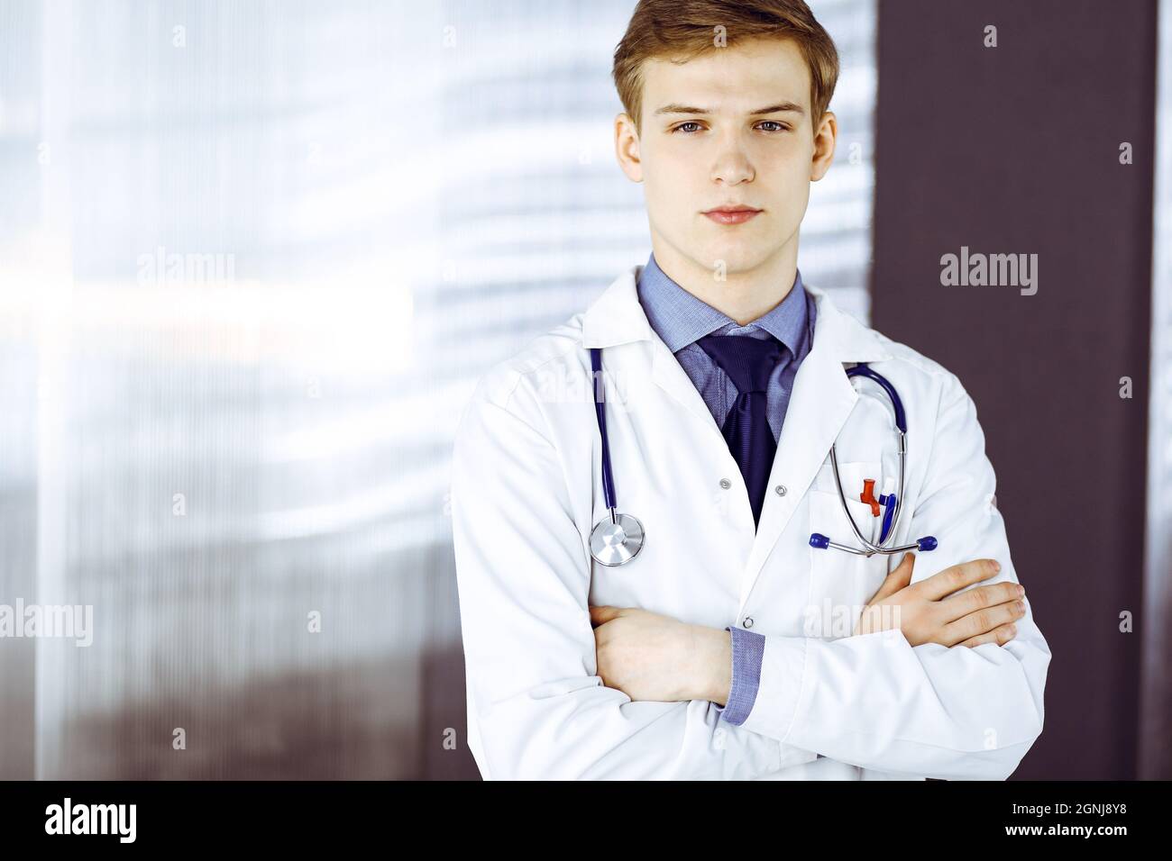 Young doctor standing with crossed arms in clinic near his working ...