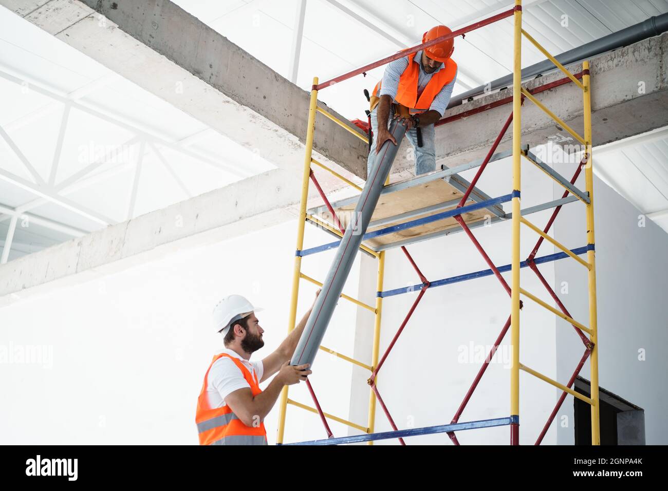 Two men builder in uniform drag the pipe on scaffolding at construction ...