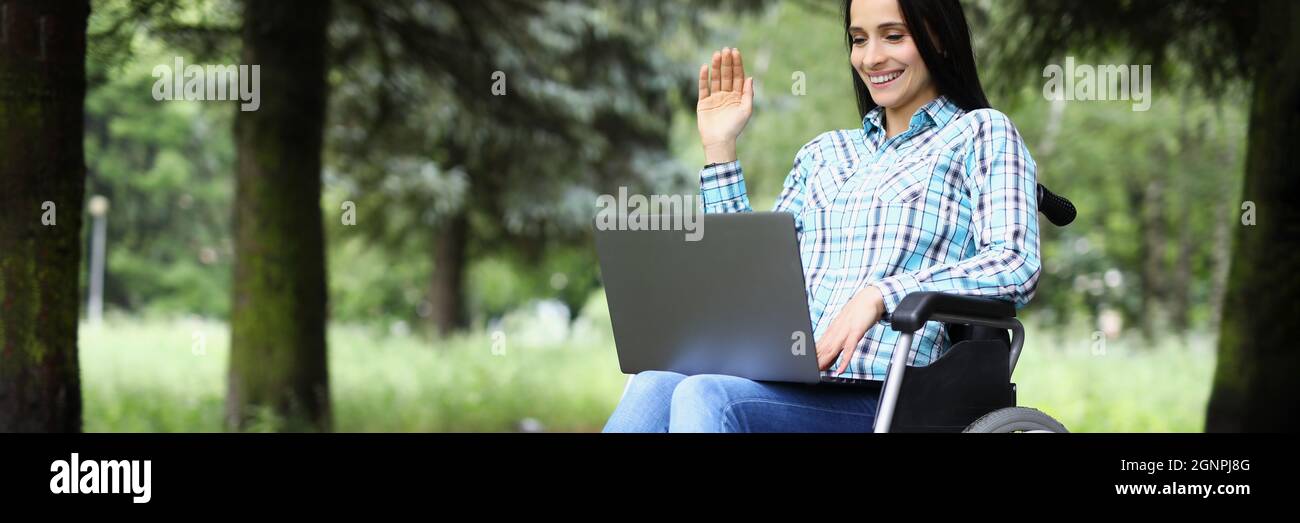 Young disabled woman in wheelchair waving hand at laptop screen in park ...