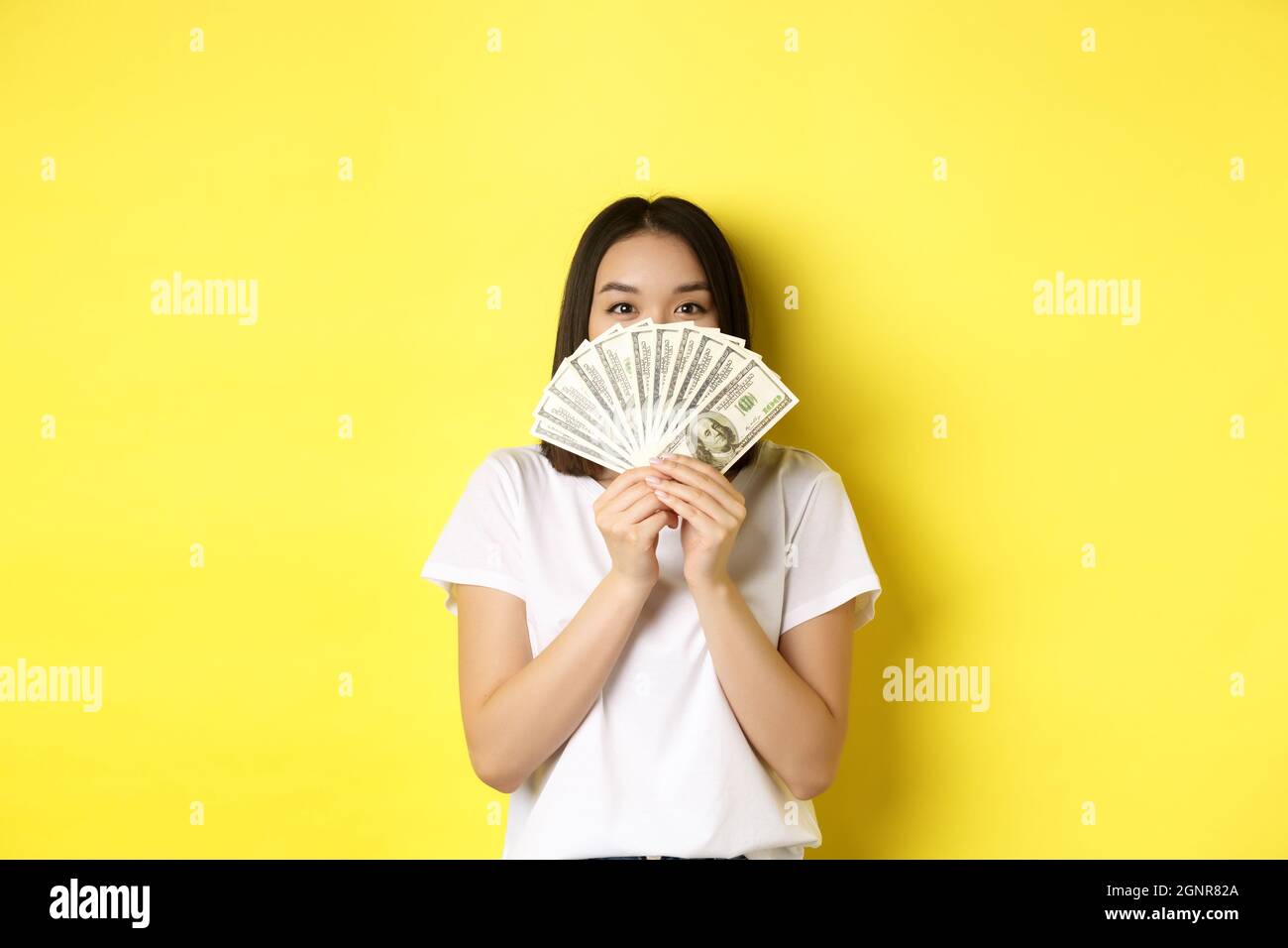 Cute asian woman hiding face behind money, peeking at camera satisfied ...