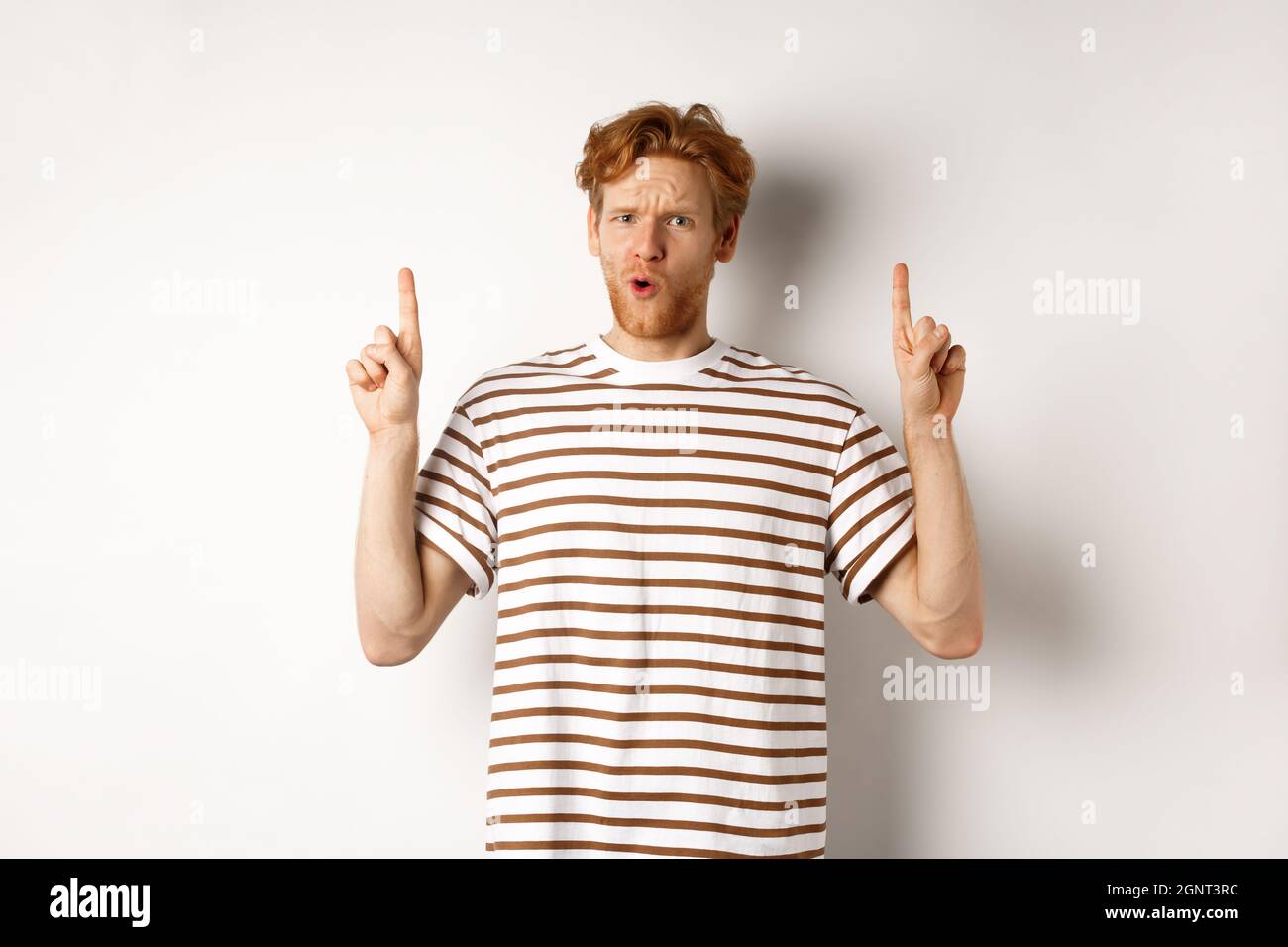 Confused young man with red curly hair pointing fingers up, staring ...