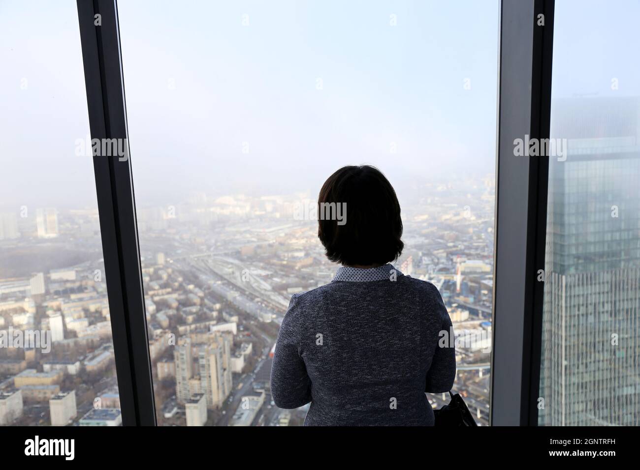 Woman standing in front of a panoramic window of skyscraper and looking ...