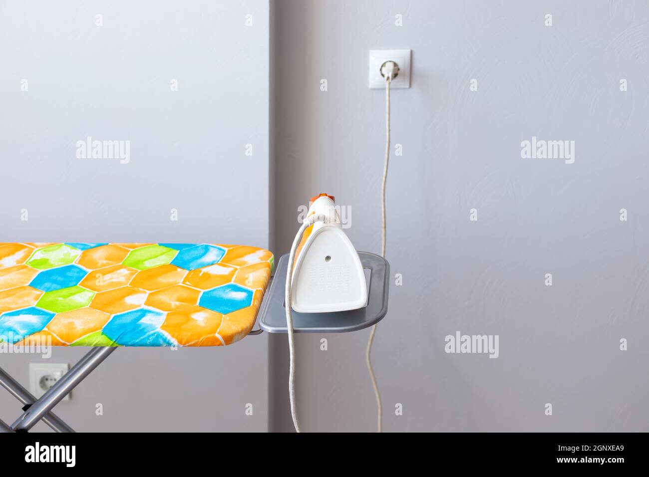 Electric iron plugged into an outlet on an ironing board against a gray ...