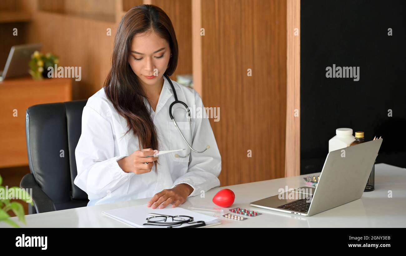 Female doctor in white gown holding or looking on clinical thermometer ...