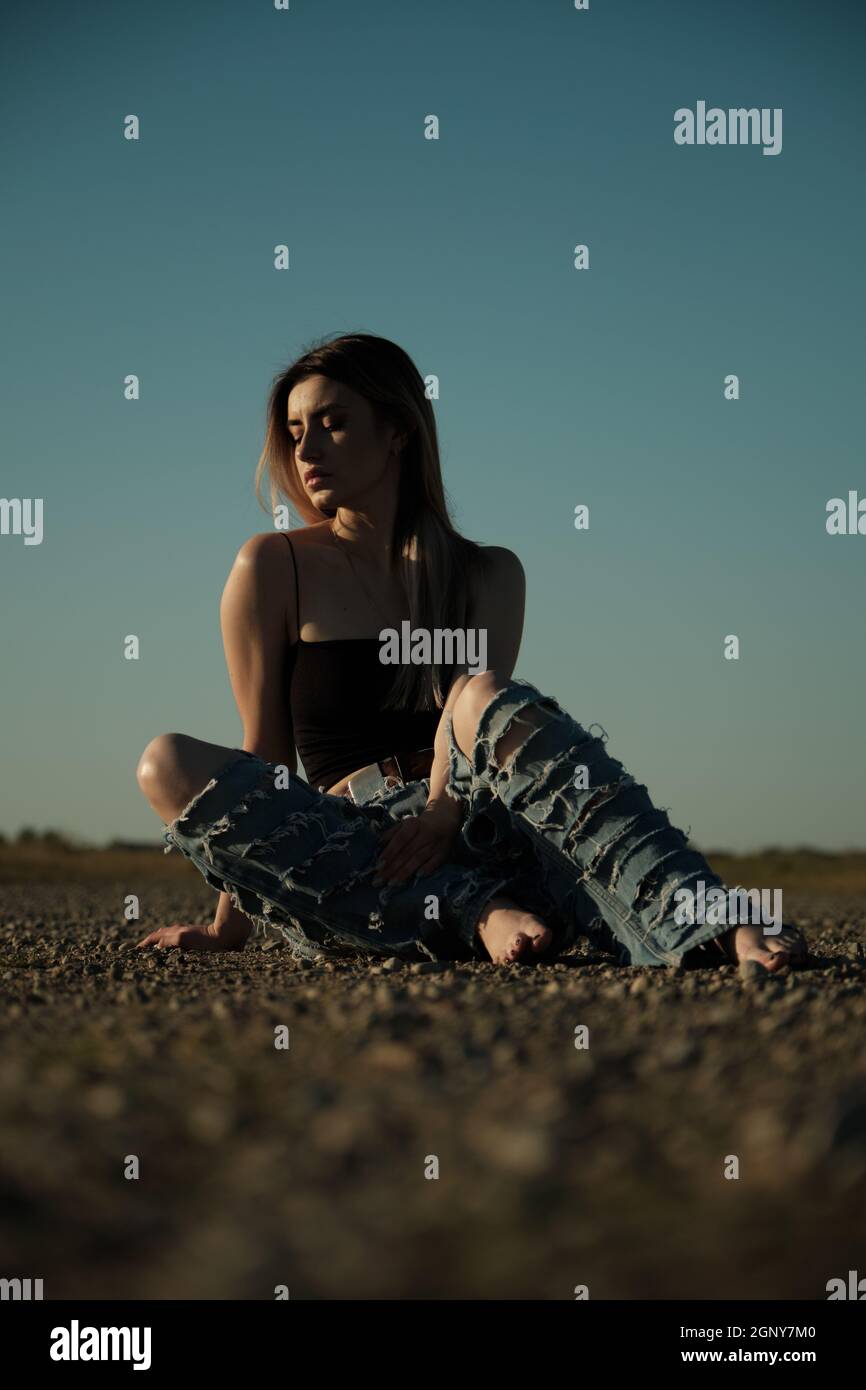 serious sad tramp girl in torn jeans sits on a deserted road Stock ...