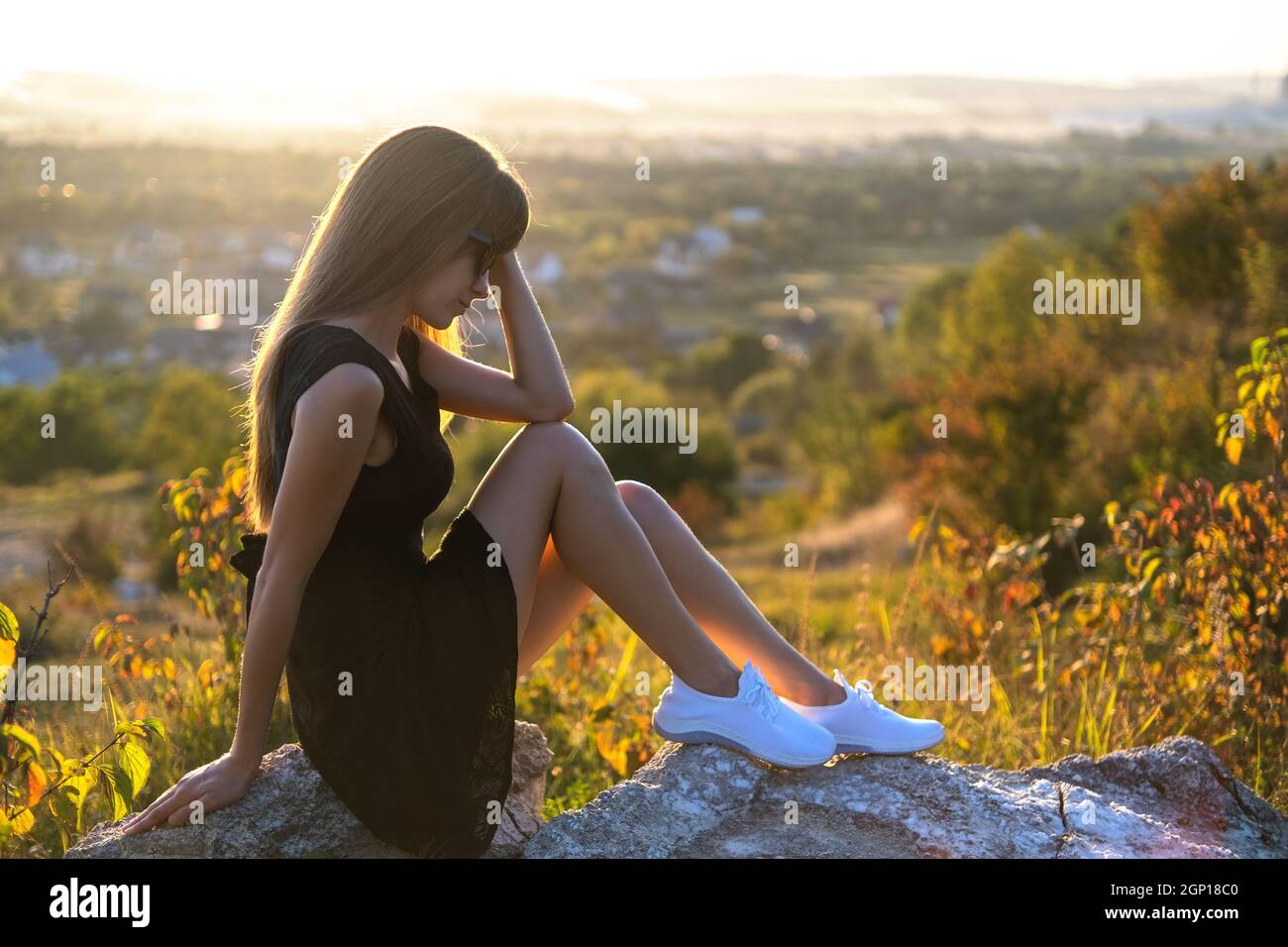 Pretty sad woman in black short summer dress sitting on a rock thinking ...