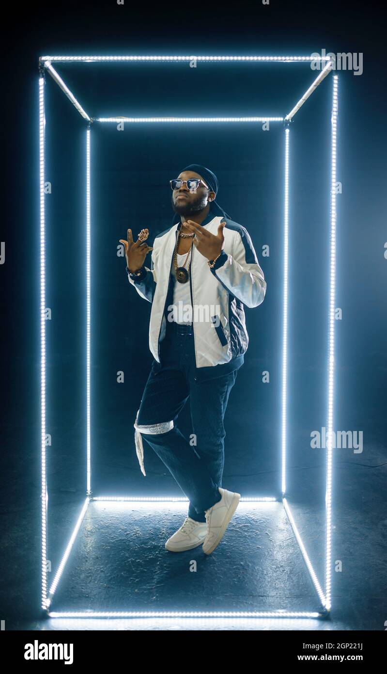 Stylish rapper in sunglasses poses in illuminated cube, dark background ...