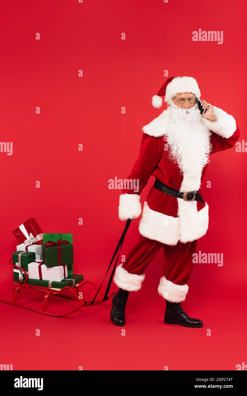 Santa talking on smartphone and pulling sleigh with presents on red ...