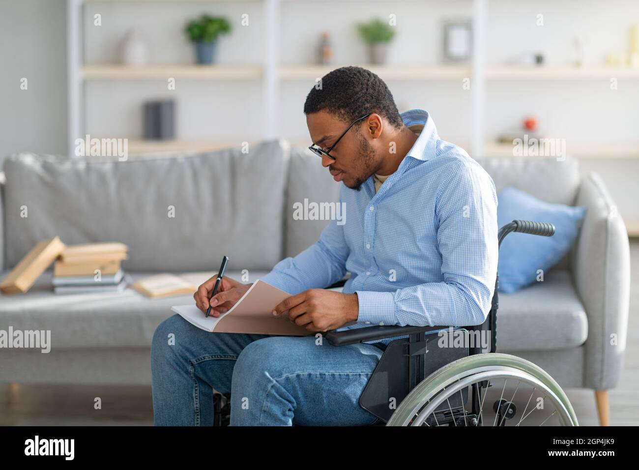 Focused handicapped black guy in wheelchair writing in notebook at home ...