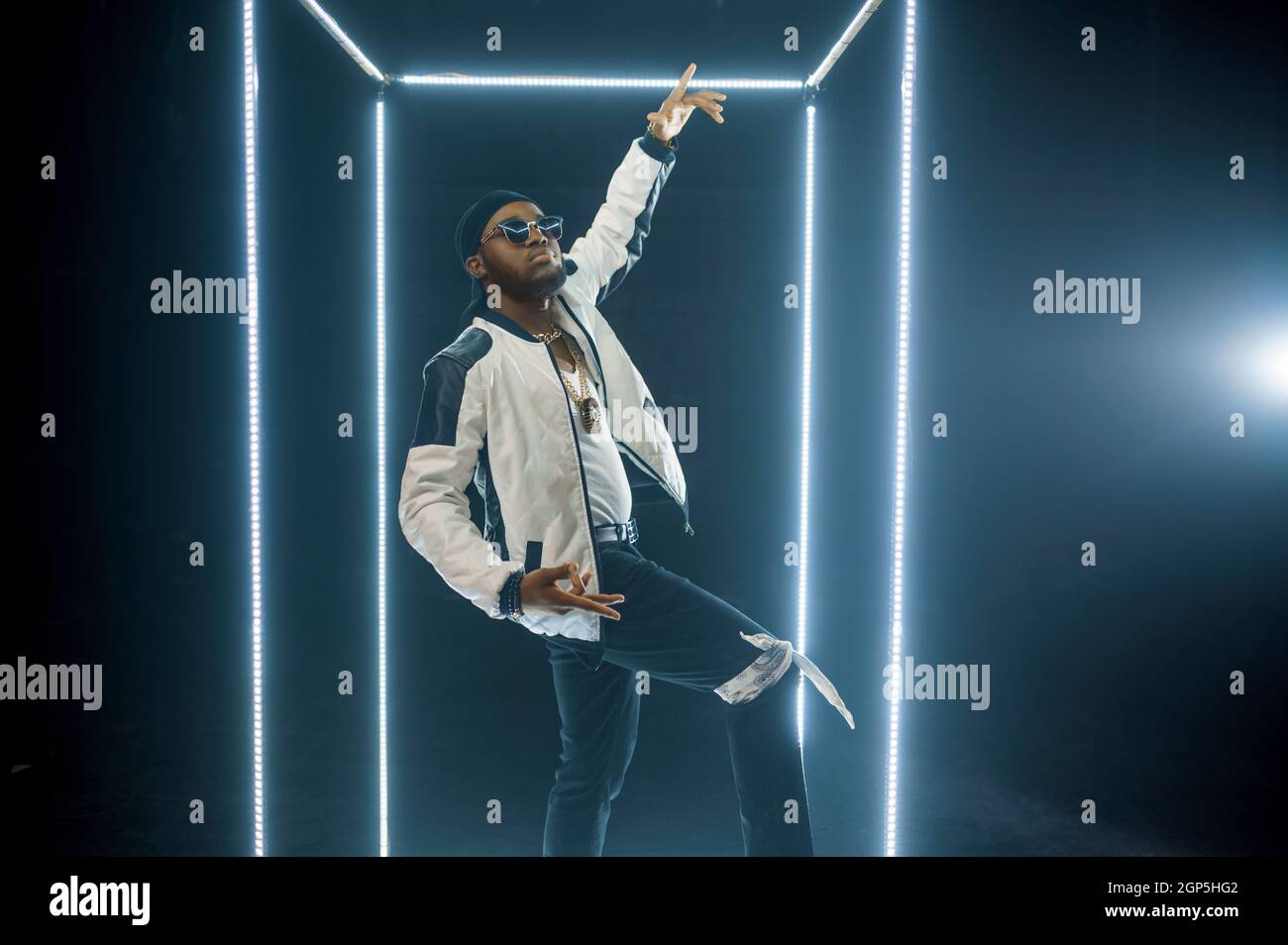 Stylish rapper in sunglasses poses in illuminated cube, dark background ...
