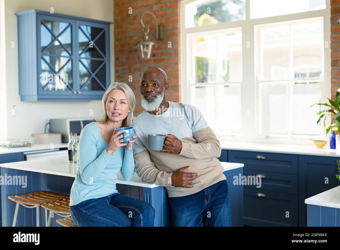 Happy senior diverse couple in kitchen sitting at countertop, drinking ...