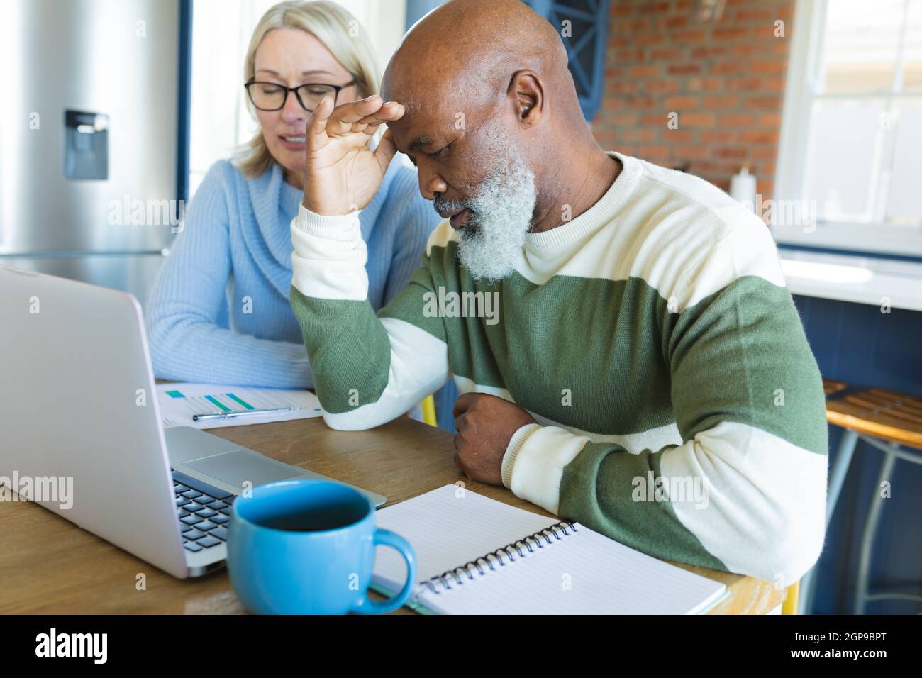 Stressed senior diverse couple in kitchen sitting at table, using ...