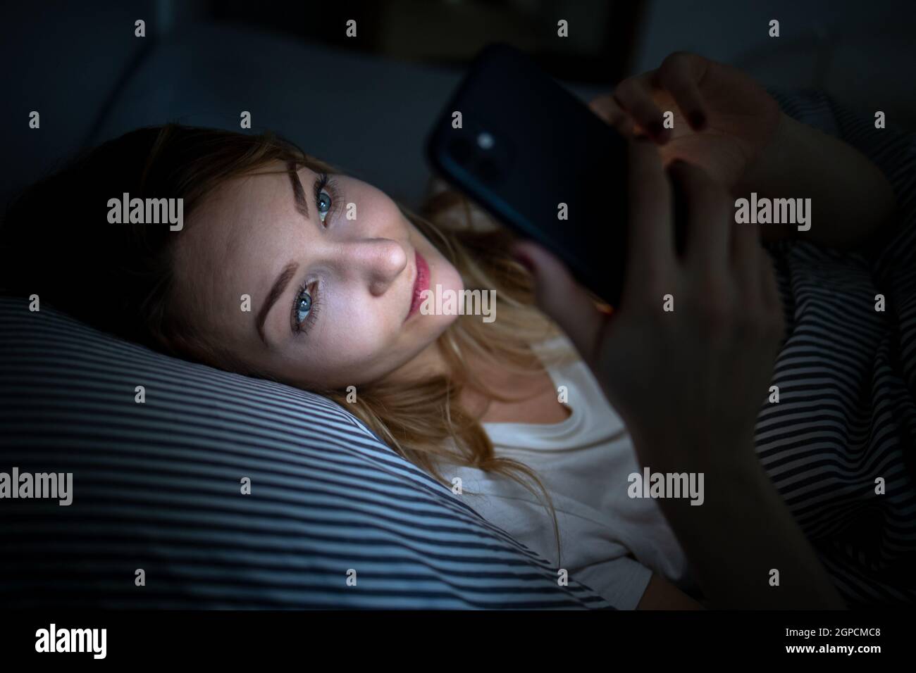 Young woman in bed holding a phone, tired and exhausted, blue light straining her eyes, messing ...