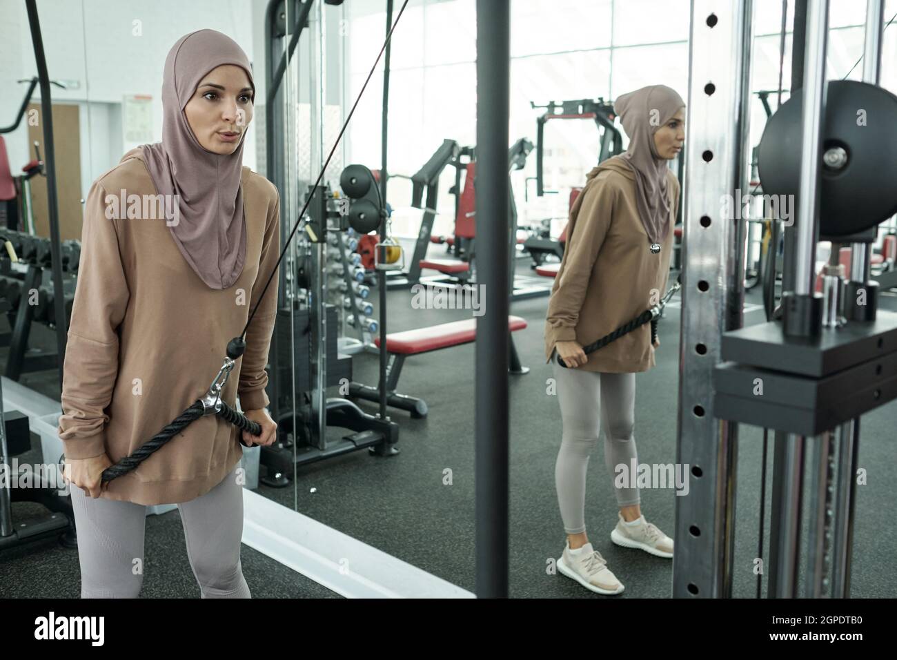 Portrait of beautiful sporty Muslim woman wearing hijab doing strengthening exercise with cables ...