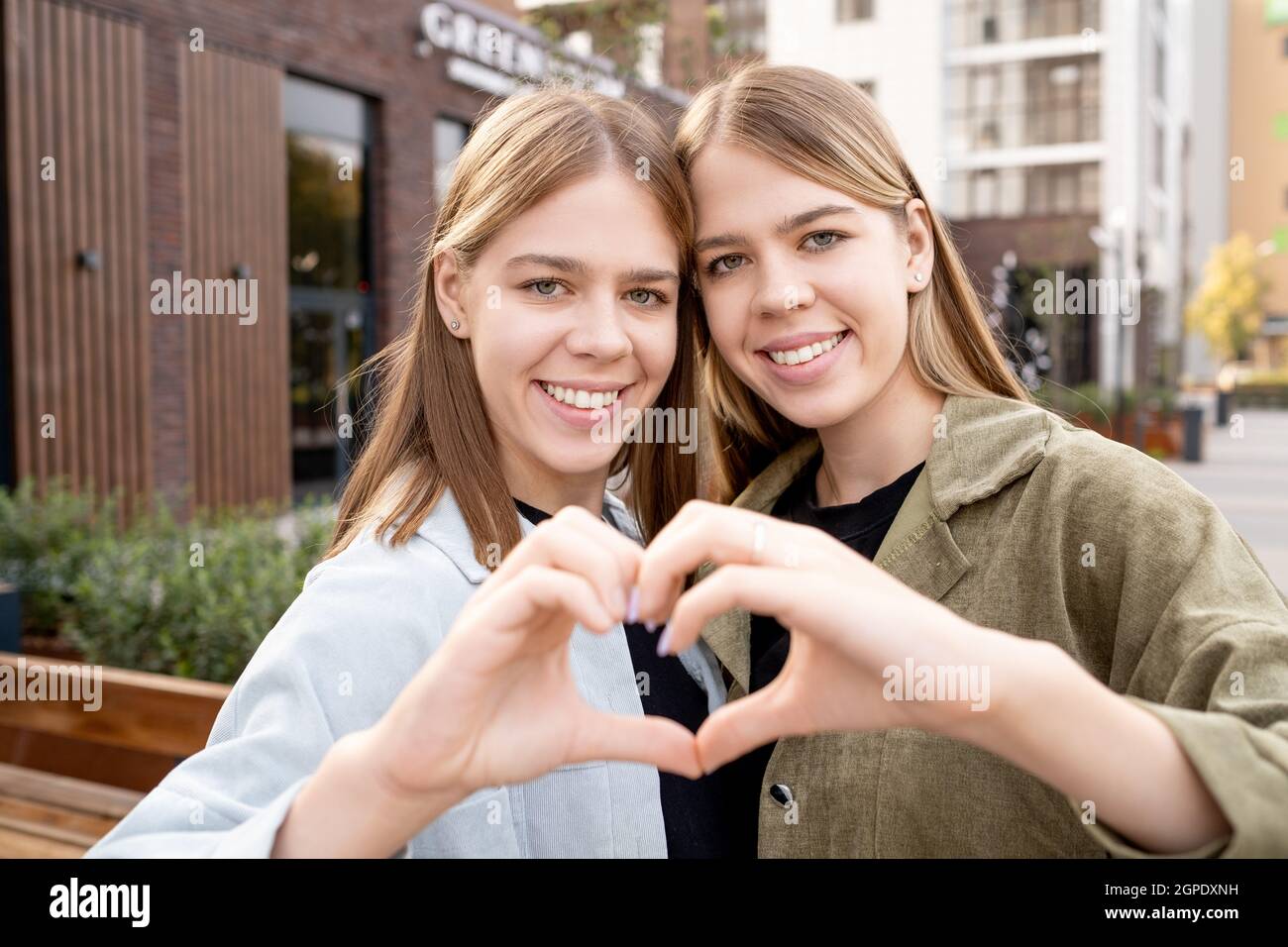 Happy affectionate twin girls in casualwear making shape of heart with ...