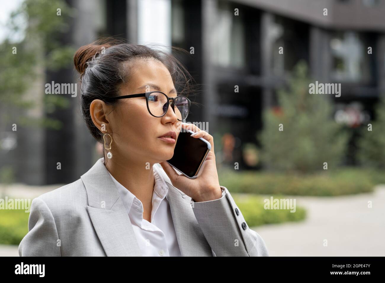 Young serious Asian businesswoman with smartphone by ear calling ...