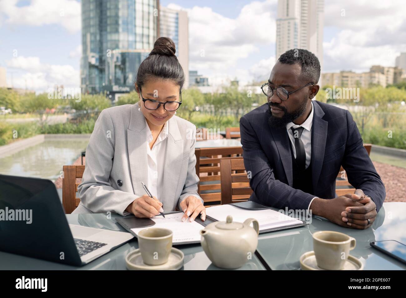 Two young elegant business partners sitting by table while one of them ...