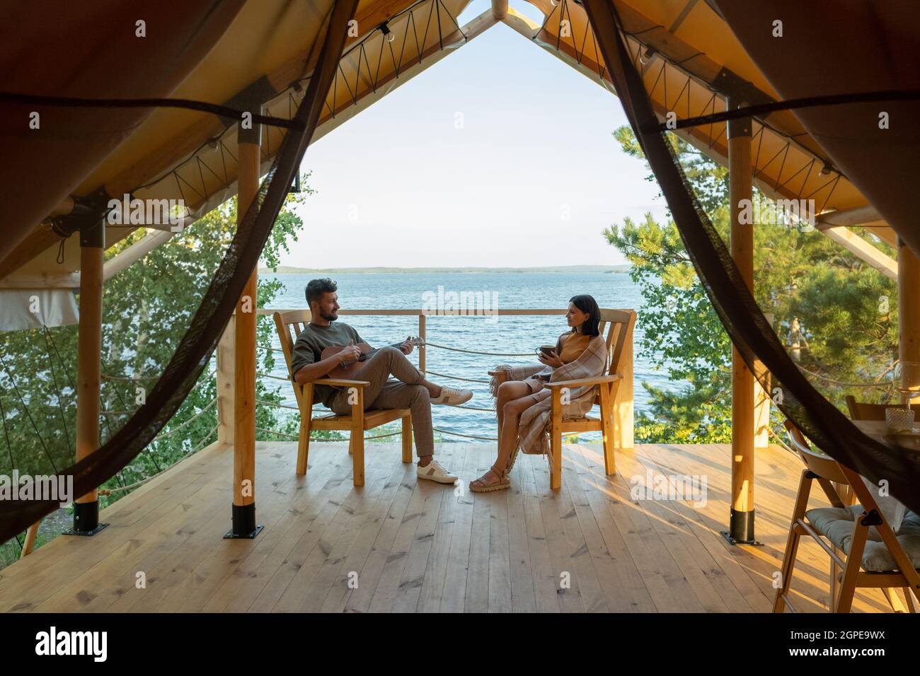 Happy amorous couple relaxing in wooden armchairs on patio by glamping ...