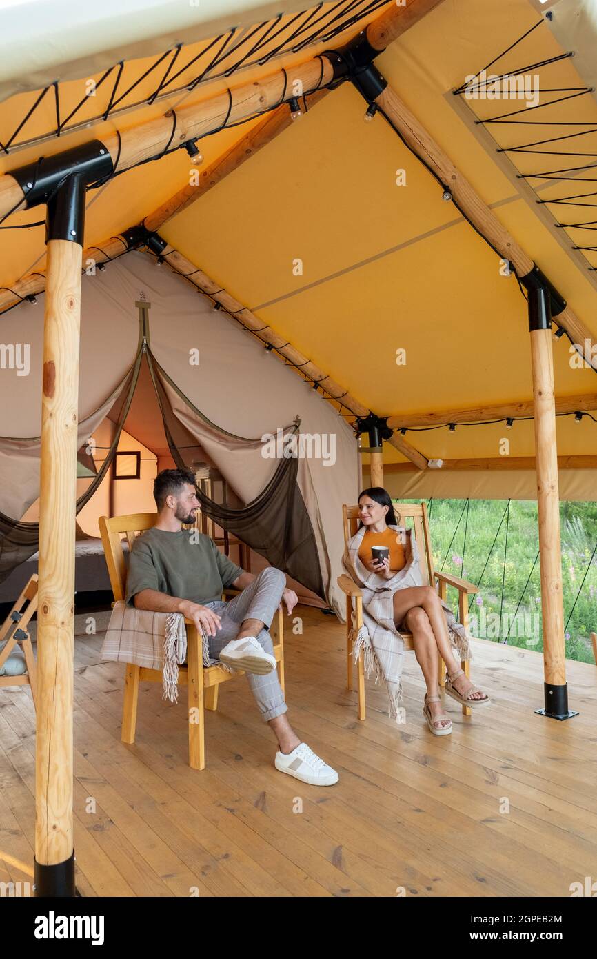 Young restful man and woman talking while relaxing in armchairs by ...