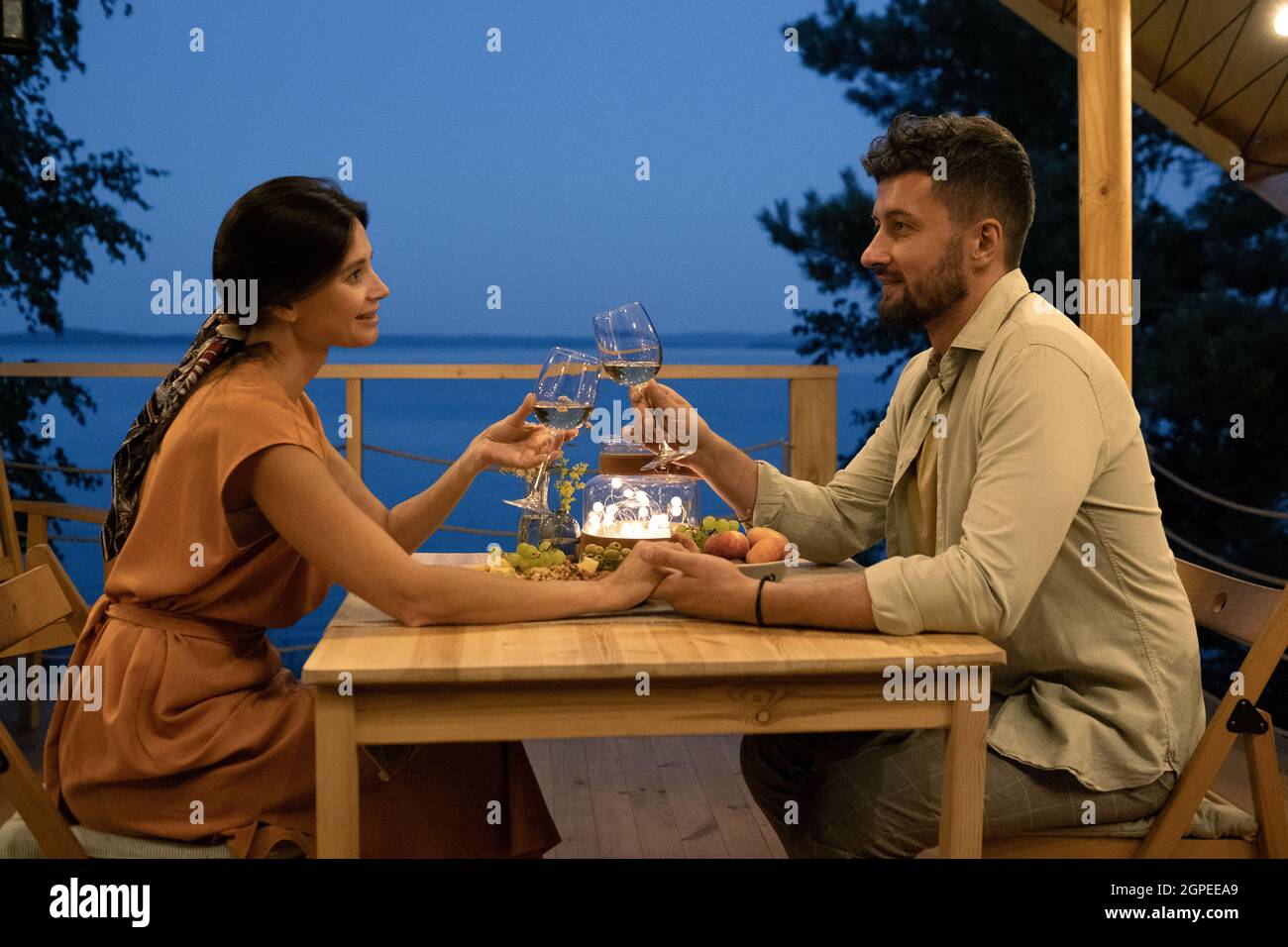 Happy couple with glasses of wine toasting by served table while ...