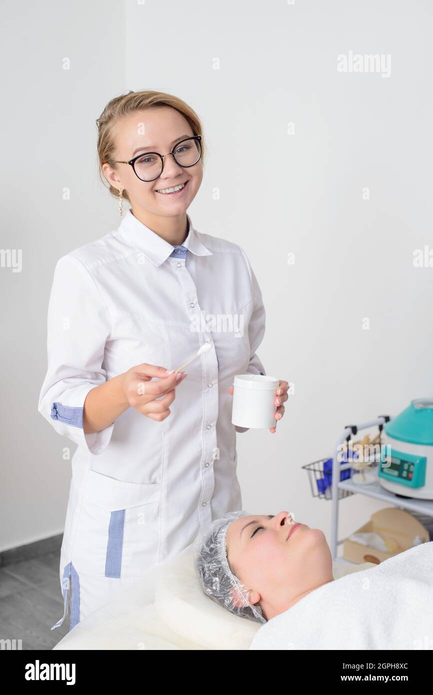 Young female cosmetologist applies cosmetic product on the face of ...