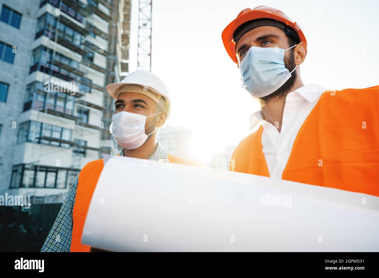 Two men in workwear and medical masks working with blueprints on object ...
