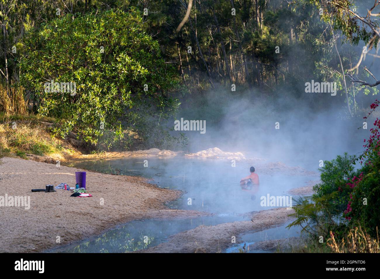 Mist rising in the early morning as man sits in shallow hot water pool ...