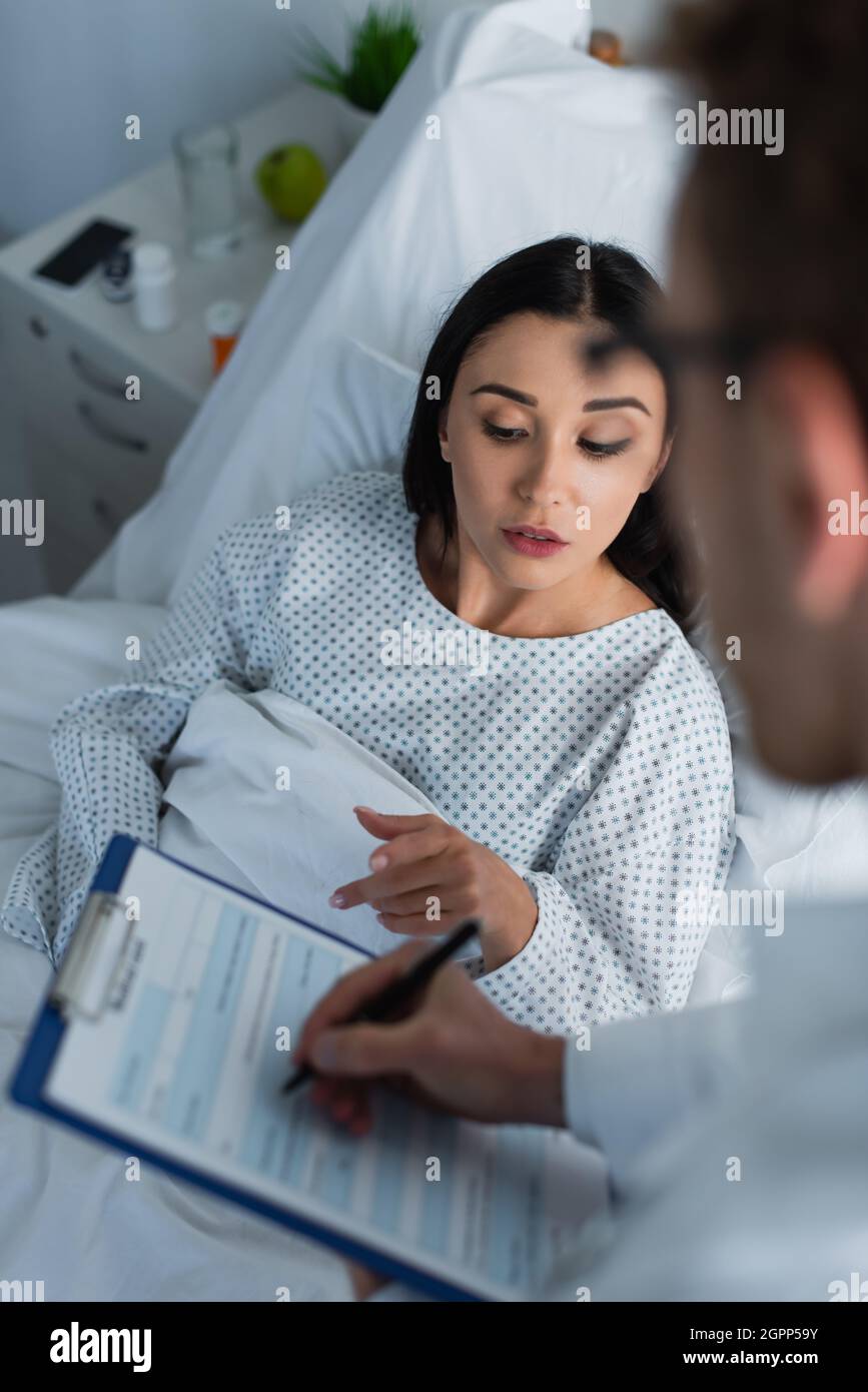 high angle view of brunette woman pointing at clipboard near blurred ...