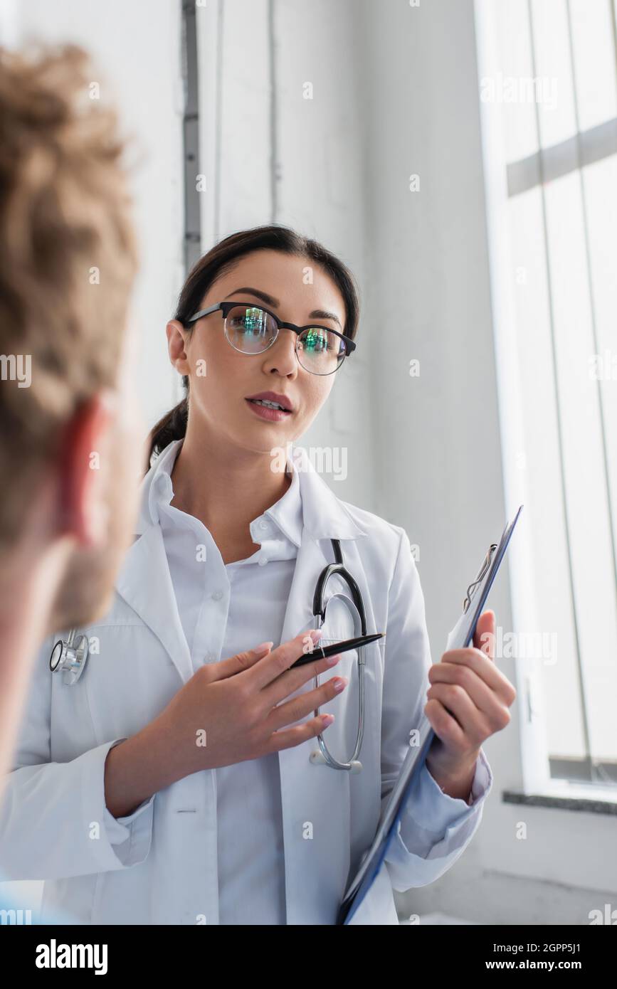 doctor in glasses holding pen and clipboard while looking at blurred ...