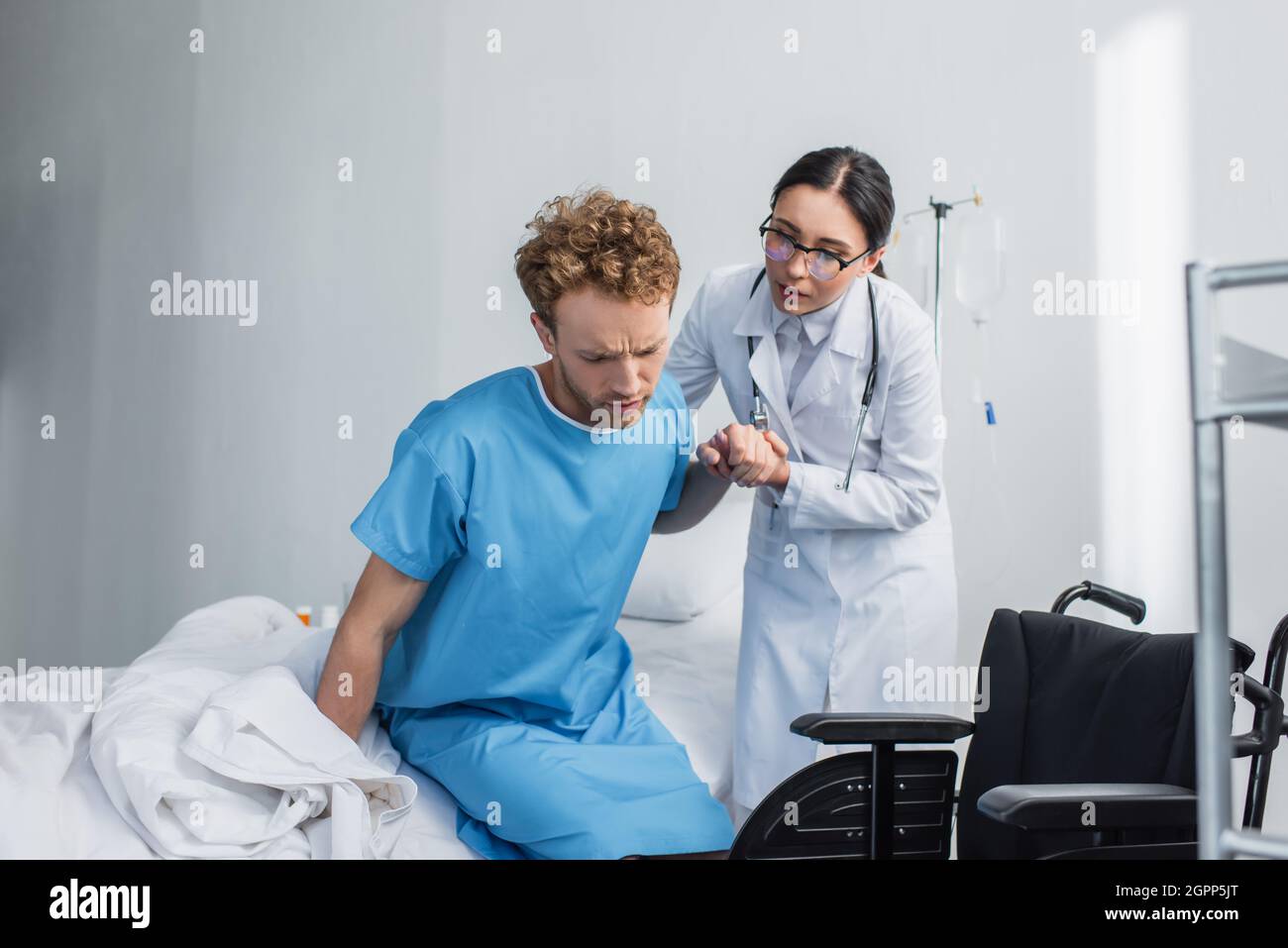 doctor in glasses helping curly patient get up from hospital bed near ...