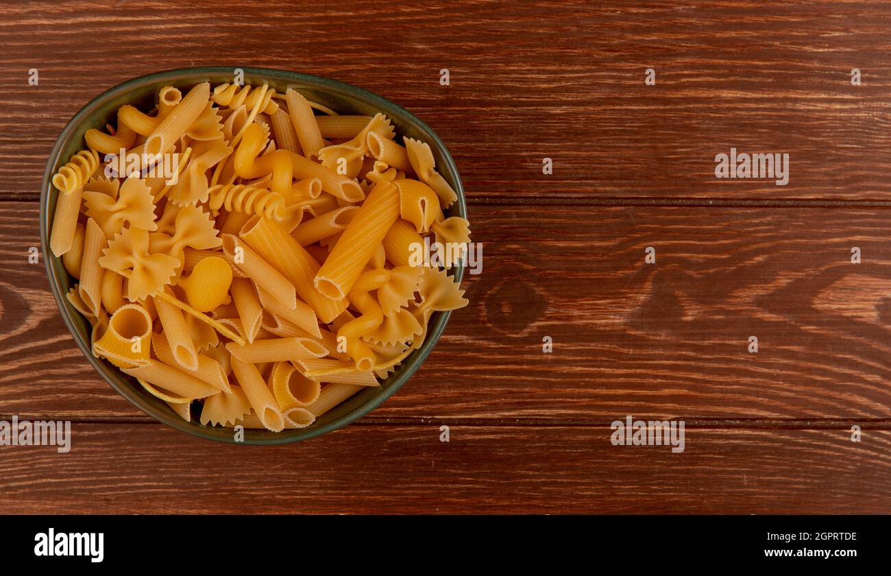 top view of different types of macaroni in bowl on wooden background ...