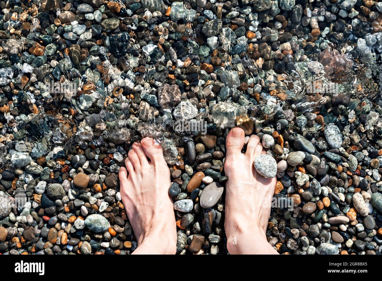 Female Feet In Sea Water On A Pebble Shore Top View In Summer Many