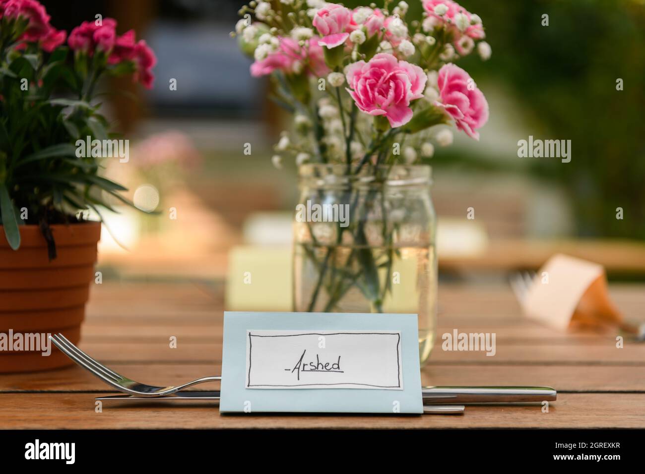 Hand written table name card and cutlery on a wooden table, flowers in ...