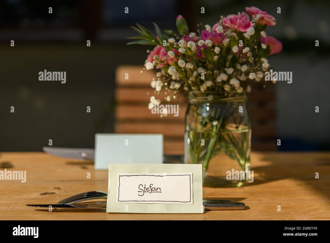 Hand written table name card and cutlery on a wooden table, flowers in ...